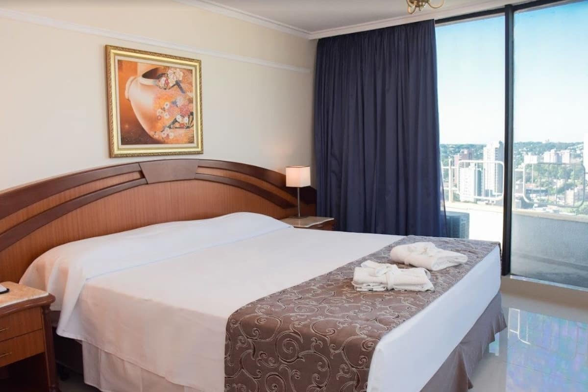 Quarto de hotel com uma cama grande, mesa de cabeceira, abajur, pintura e vista da cidade através de grandes janelas.