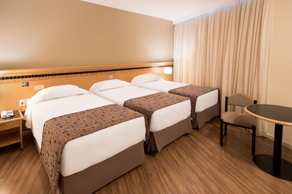 Quarto de hotel com duas camas, uma mesa pequena, uma cadeira e uma mesinha de cabeceira com telefone.