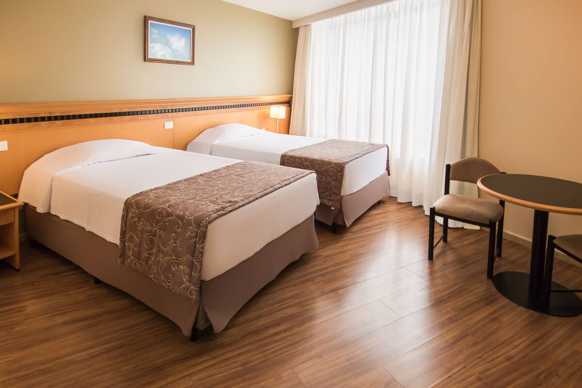Quarto de hotel com duas camas, uma mesinha e uma cadeira.