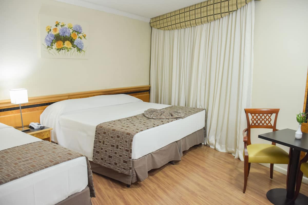 Quarto de hotel com duas camas, uma cabeceira de madeira, uma mesa de jantar com uma cadeira e um quadro na parede.
