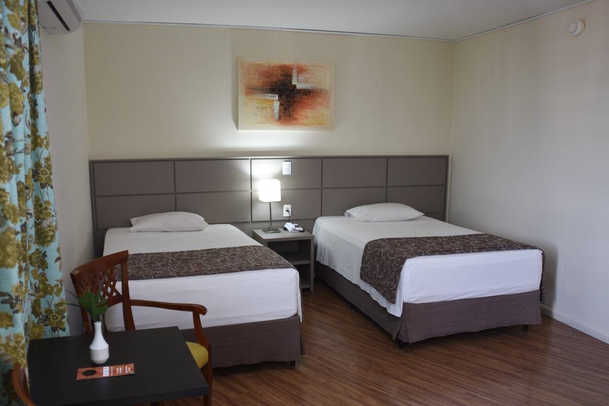 Quarto de hotel com duas camas de solteiro, uma mesinha com uma cadeira, mesa de cabeceira com abajur e uma obra de arte na parede.