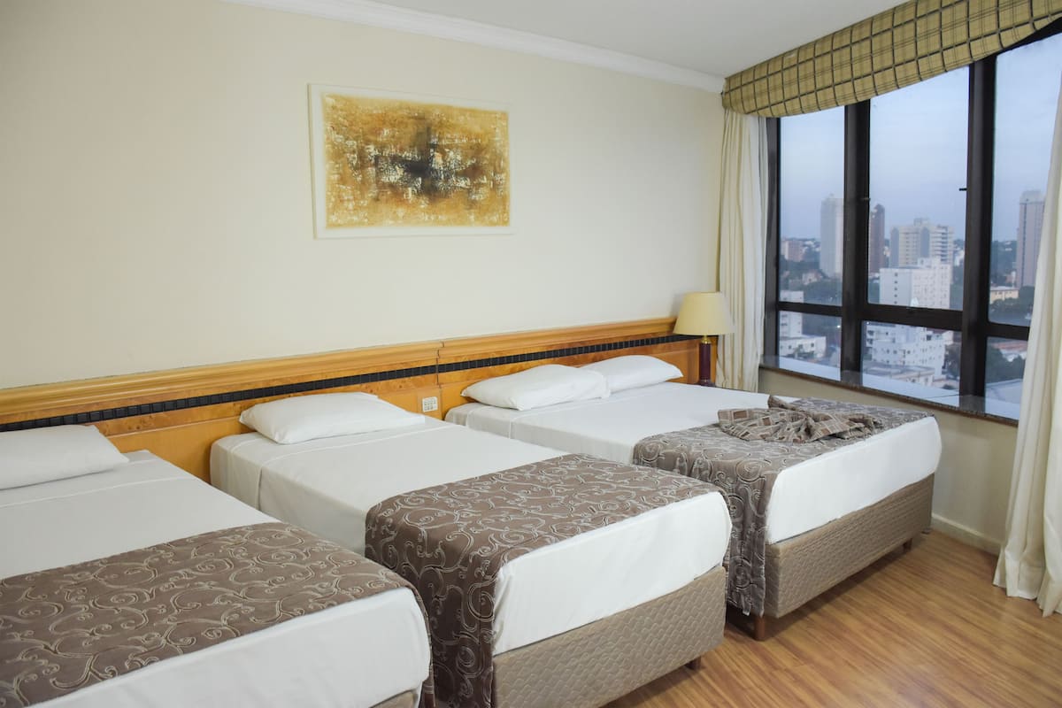 Quarto de hotel com três camas, uma janela grande com vista para a cidade e um quadro na parede.