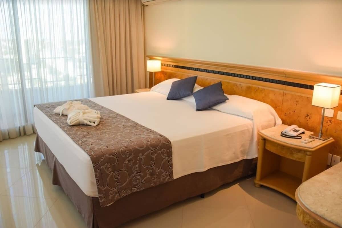 Quarto de hotel com uma cama grande, uma mesa de cabeceira com abajur e um telefone.