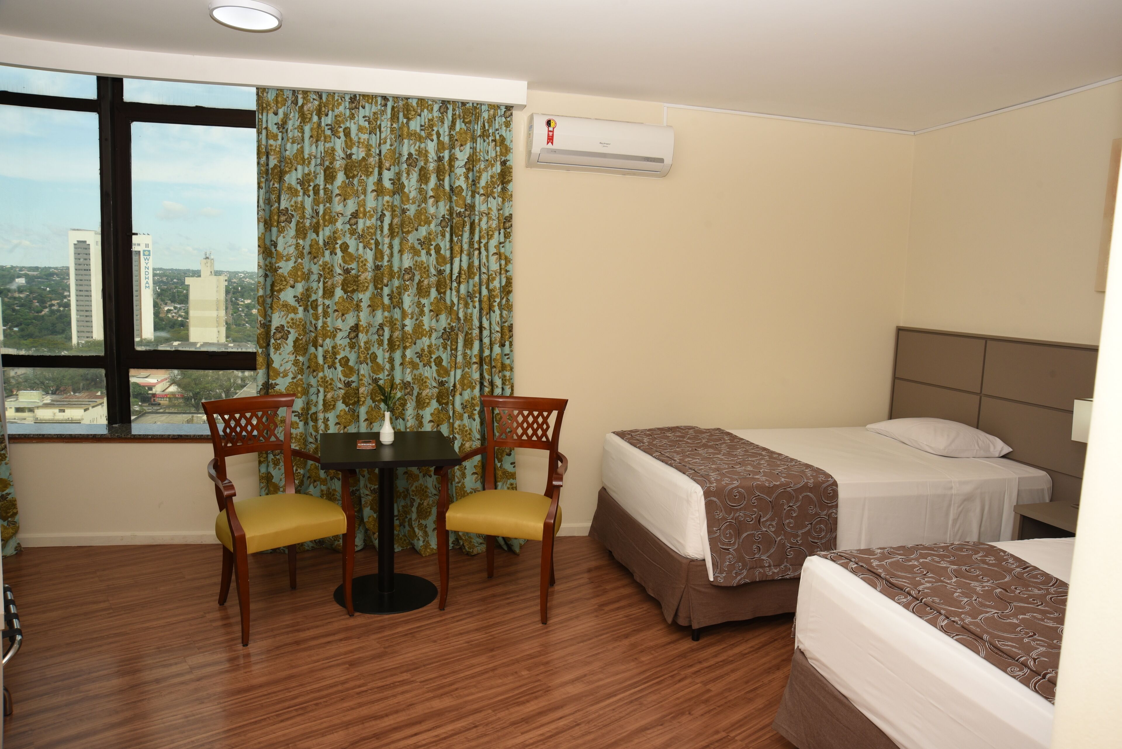 Quarto de hotel com duas camas, uma mesinha com cadeiras, uma janela com cortinas e um aparelho de ar condicionado.