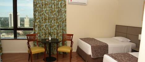 Quarto de hotel com duas camas, uma mesinha com cadeiras, uma janela com cortinas e um aparelho de ar condicionado.