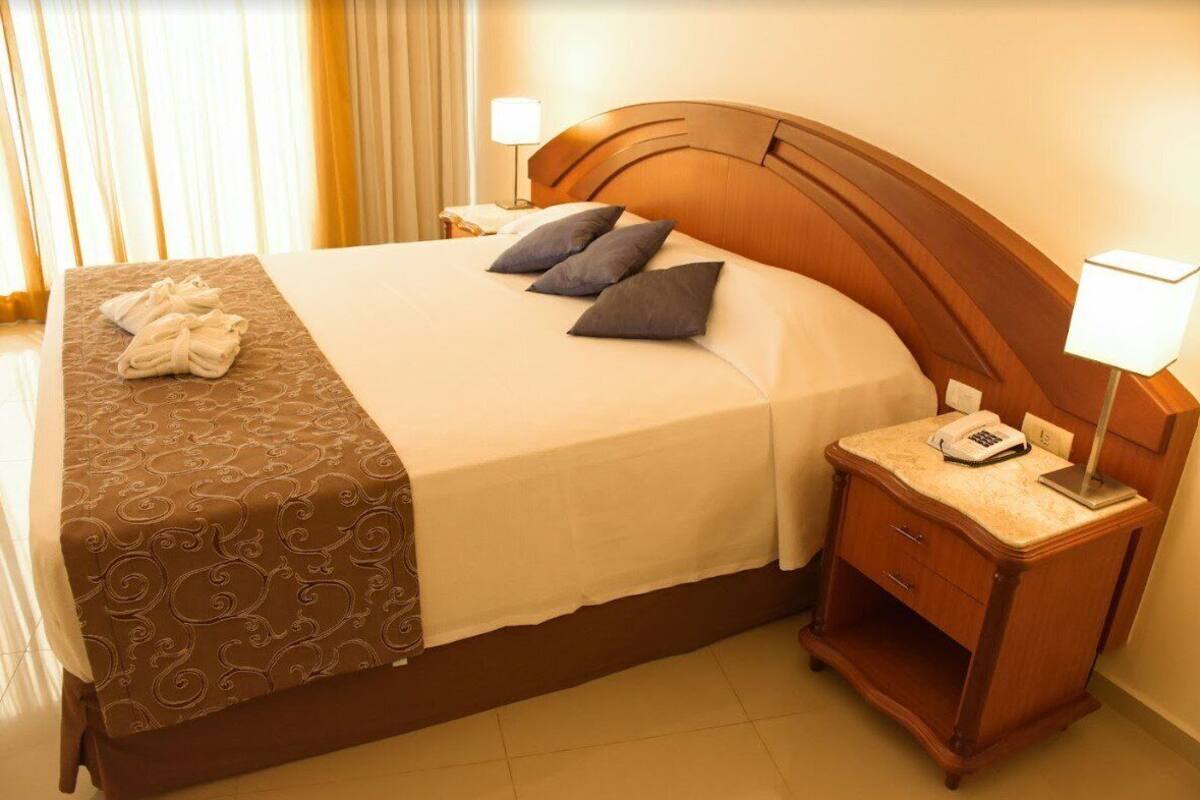 Quarto de hotel com uma cama grande, uma mesa de cabeceira com abajur e um telefone.