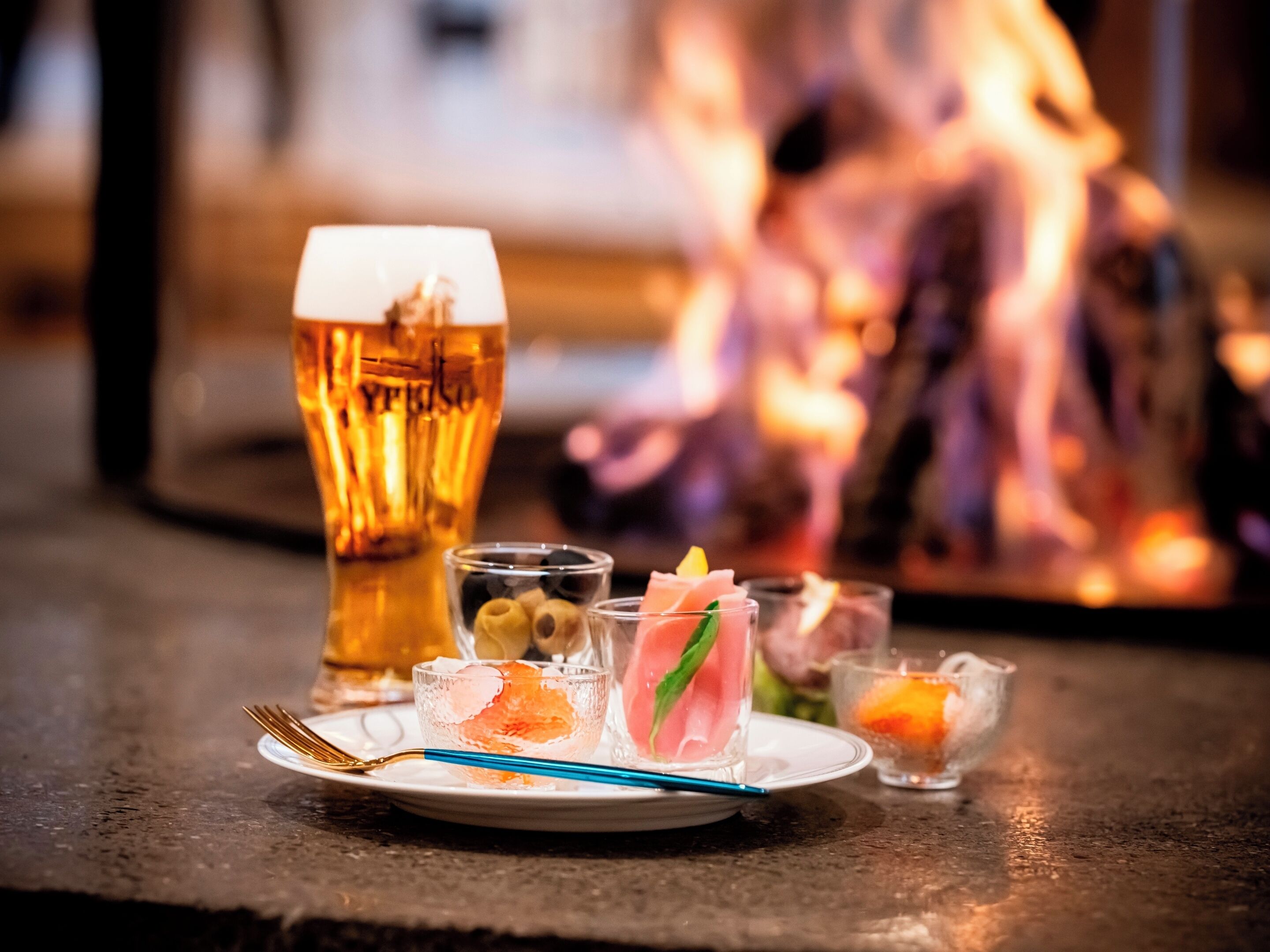 Uma mesa de jantar com diversos petiscos e bebidas, incluindo um copo grande de cerveja, um copo de coquetel e pequenos copos para shots com guarnições.