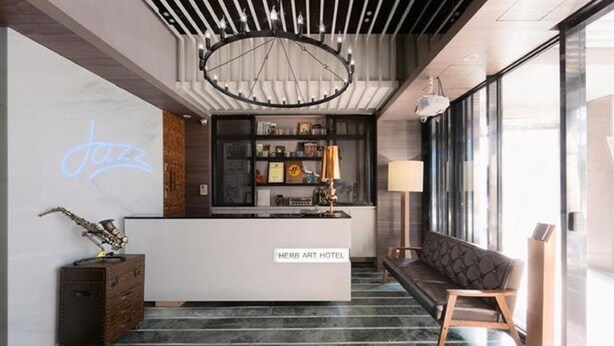 Um lobby com uma mesa de recepção identificada como "HERB ART HOTEL", um sofá, um abajur e uma parede com um letreiro de neon.