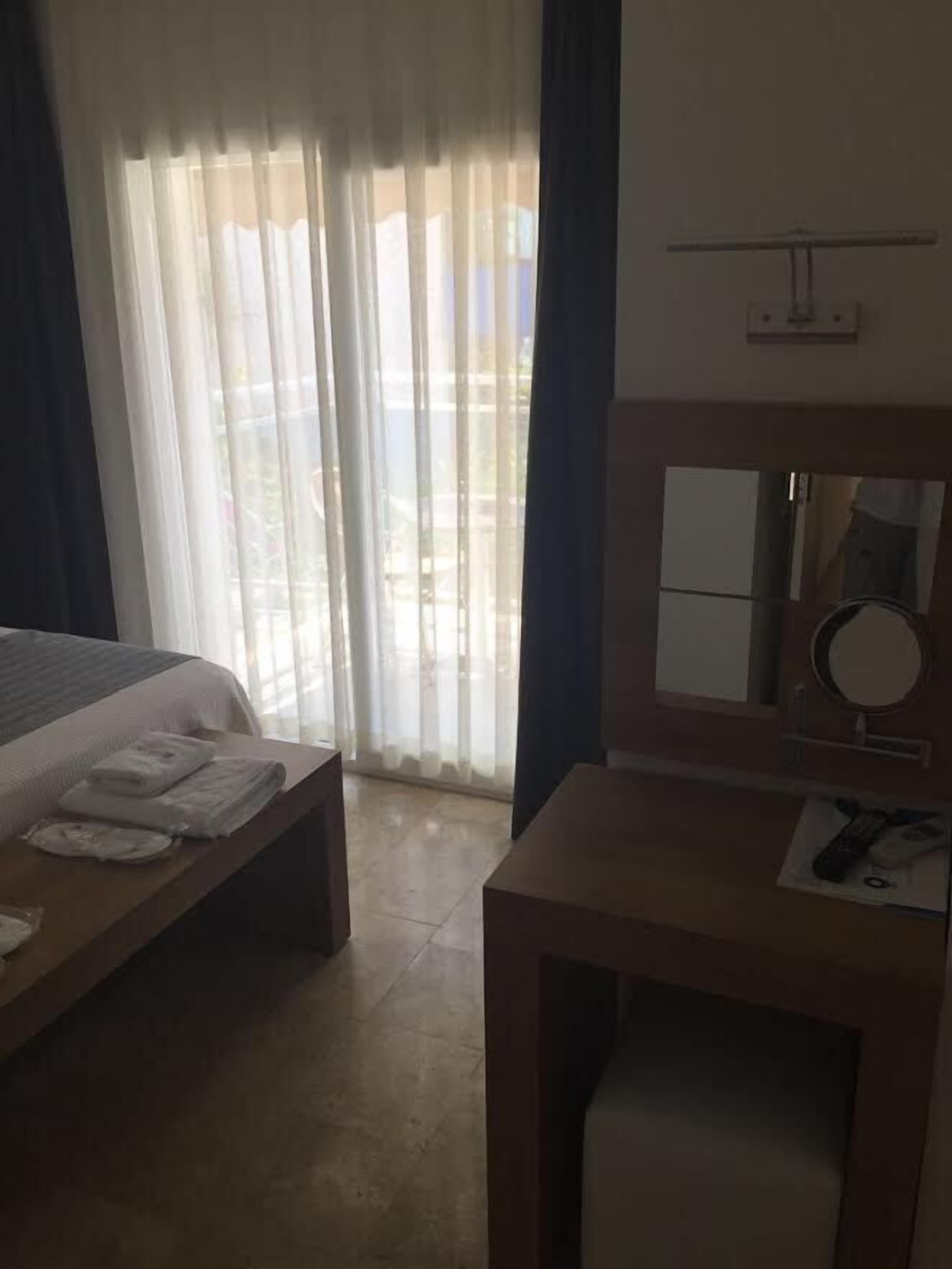 Quarto de hotel com cama, mesa de cabeceira com espelho e janela com cortinas translúcidas.