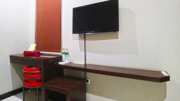 Um quarto pequeno com uma TV fixada na parede, uma escrivaninha de madeira com uma cadeira e um banquinho.