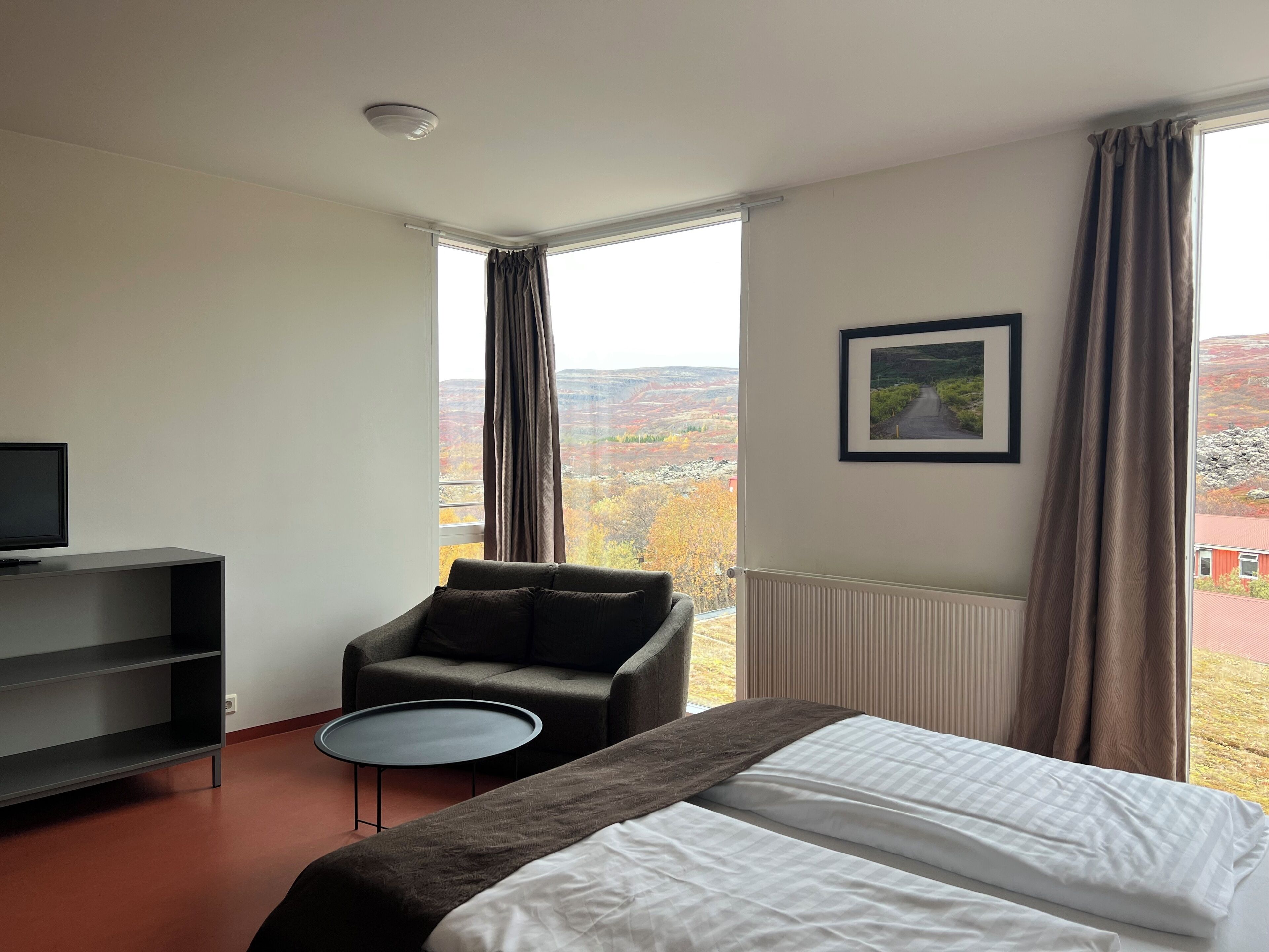 Quarto de hotel com cama, uma cadeira, mesa pequena, televisão e janela com vista para a paisagem.