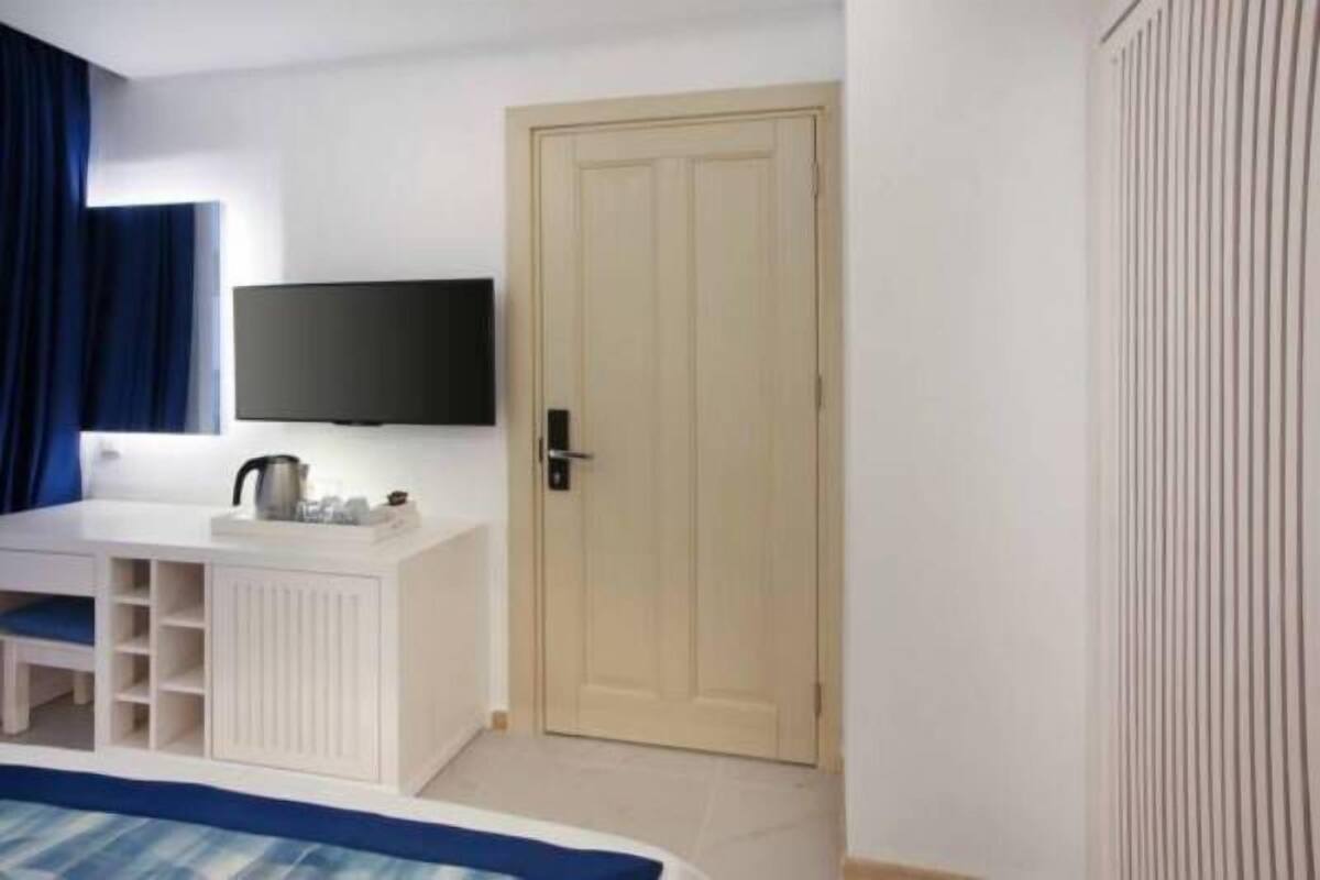 Quarto com cama, mesa de cabeceira branca com chaleira, televisão presa na parede e porta de madeira.
