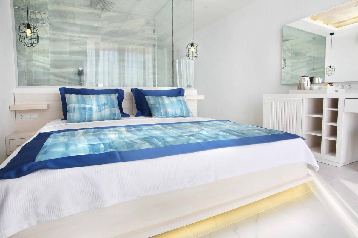 Quarto moderno com cama grande, box de vidro e cômoda branca.