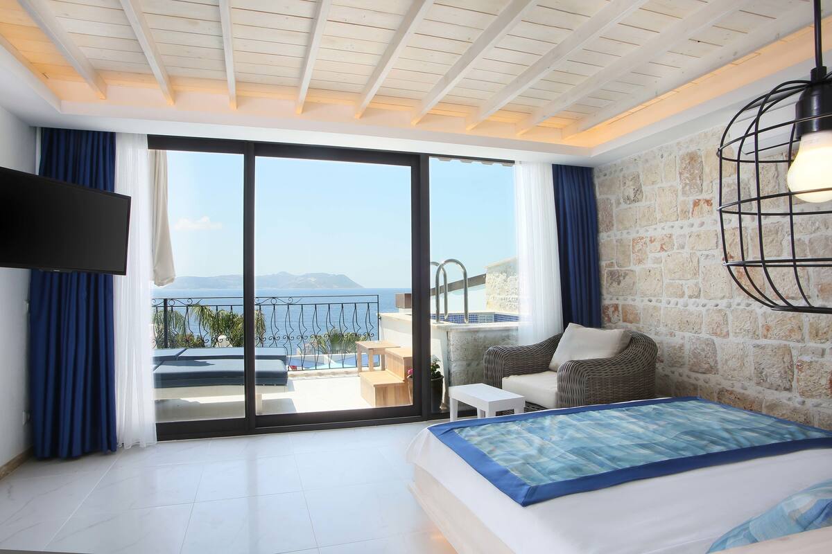 Quarto com cama, televisão, varanda e vista para o mar, além de uma parede de pedra.