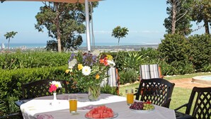 Uma área de refeições ao ar livre com uma mesa posta para dois, um vaso de flores e vista para a cidade e oceano.