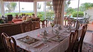 Uma área de refeições com uma mesa posta para chá, rodeada por cadeiras, com vista para vegetação e uma piscina ao fundo.