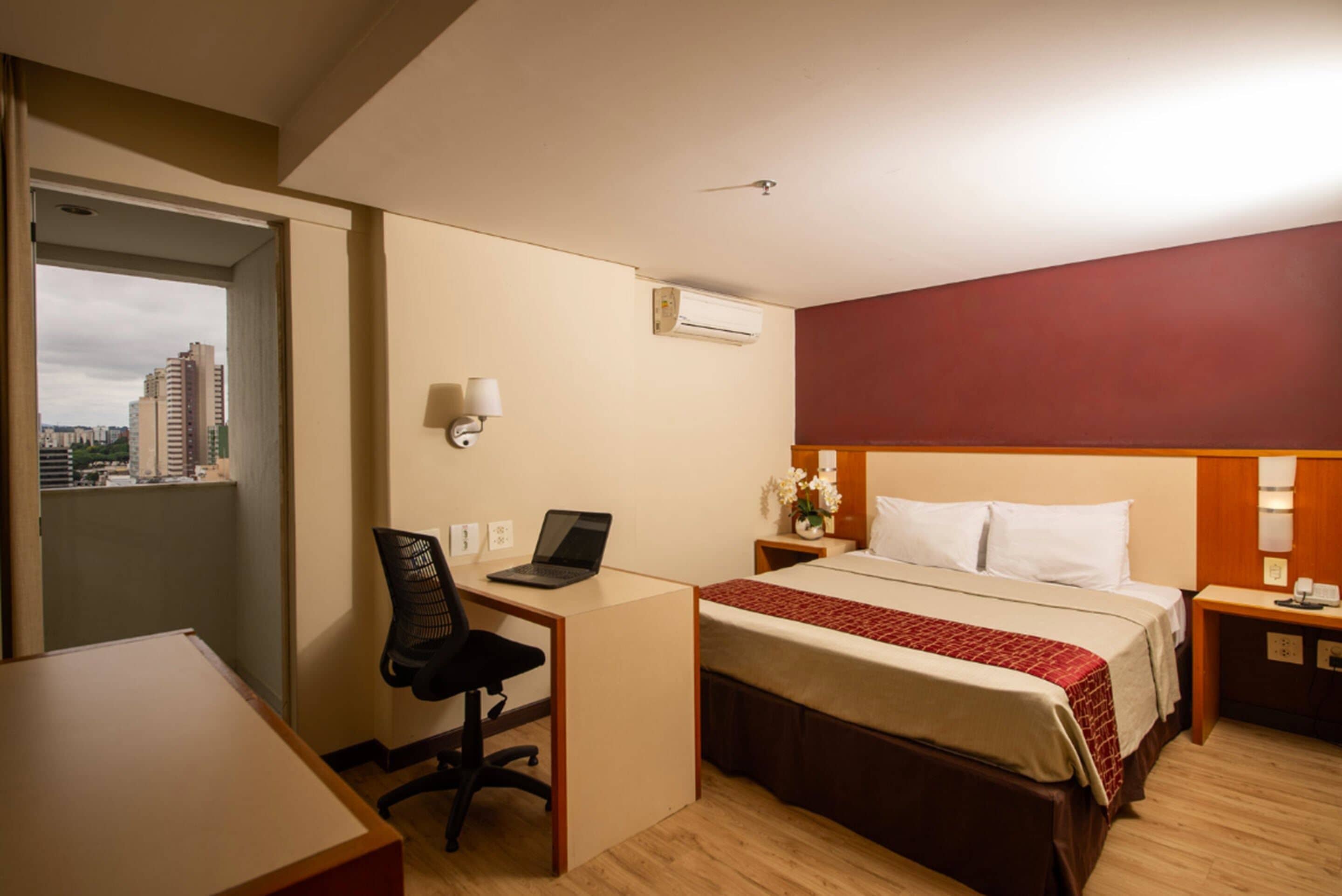 Quarto de hotel com cama, escrivaninha, cadeira e vista para a cidade.