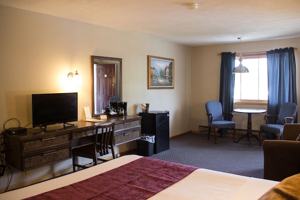 Quarto de hotel com cama, escrivaninha com computador, cadeira, mesinha e janela com cortinas.