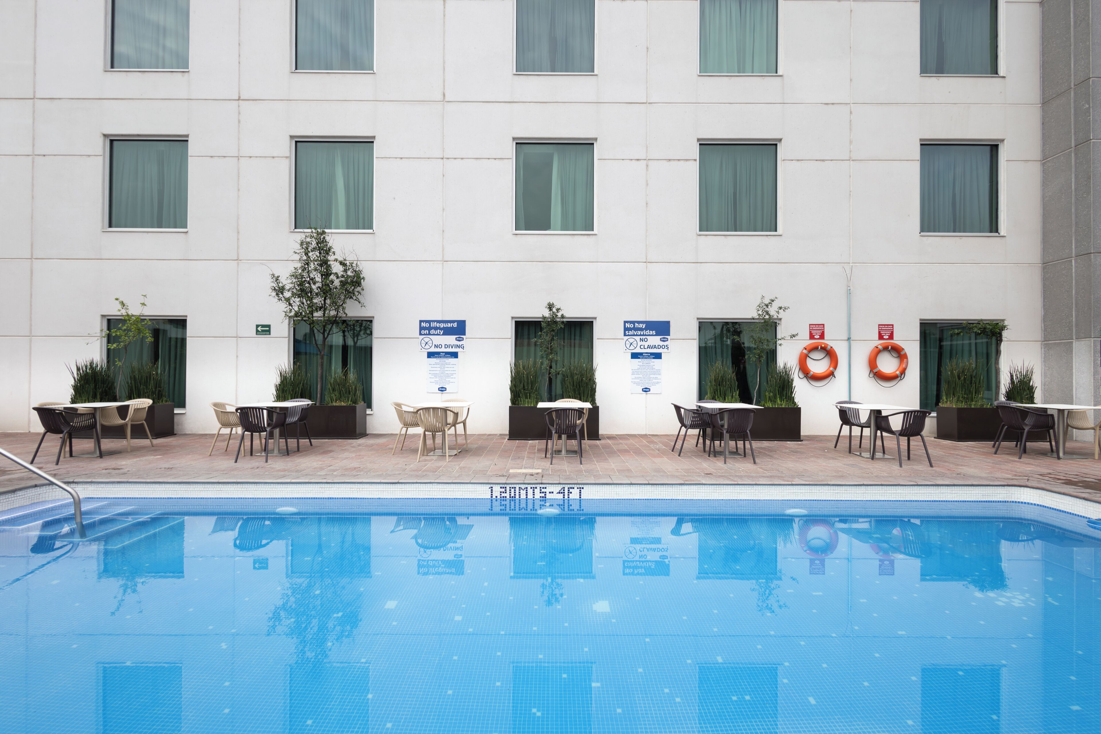 Piscina com espreguiçadeiras e mesas, um edifício com janelas e placas, e equipamentos de salvamento.
