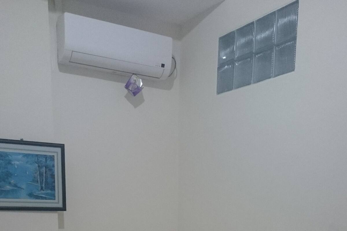 Quarto com cama, roupa de cama branca, manta vermelha, travesseiro branco e cabeceira branca. Ar condicionado fixado na parede, janela com persianas e quadro emoldurado na parede.