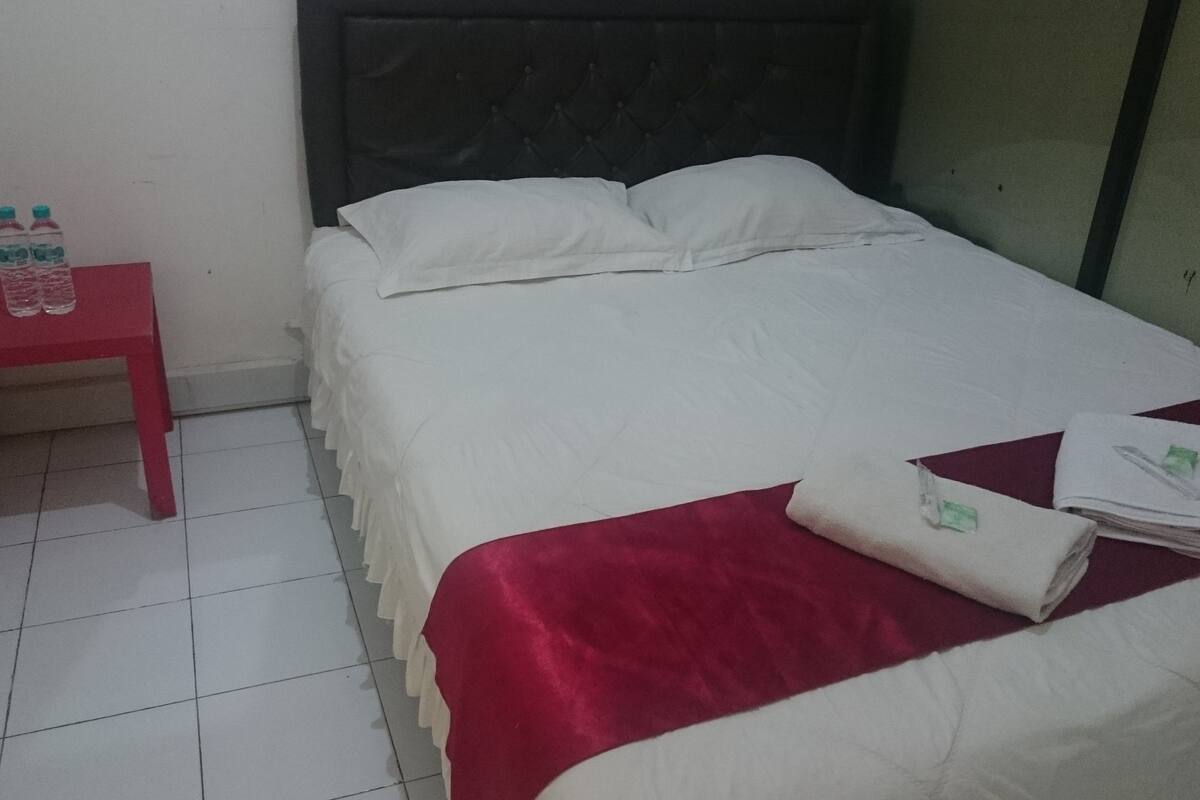 Quarto de hotel com uma cama, uma mesinha de cabeceira vermelha com duas garrafas de água e uma toalha dobrada na cama.