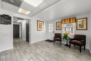 Um saguão de hotel com piso de madeira, duas poltronas, uma planta em vaso e obras de arte emolduradas nas paredes.