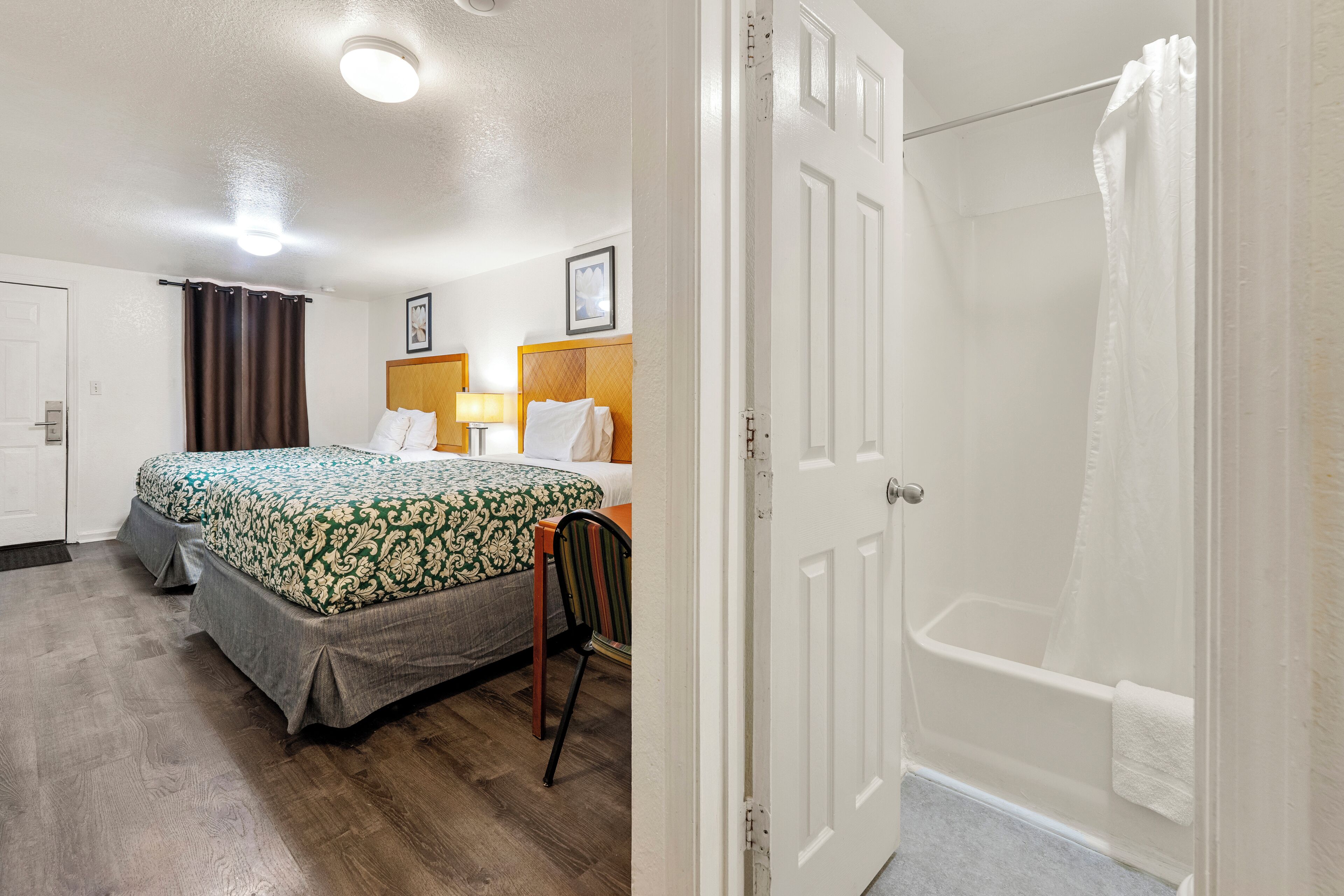 Quarto de hotel com cama, cadeira, mesa de cabeceira, luminária, cortina do chuveiro e porta.