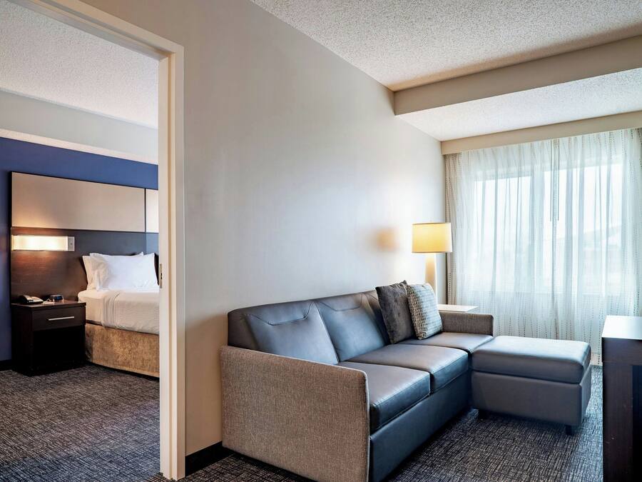 Quarto de hotel com cama, sofá, escrivaninha, abajur e janela com cortinas.