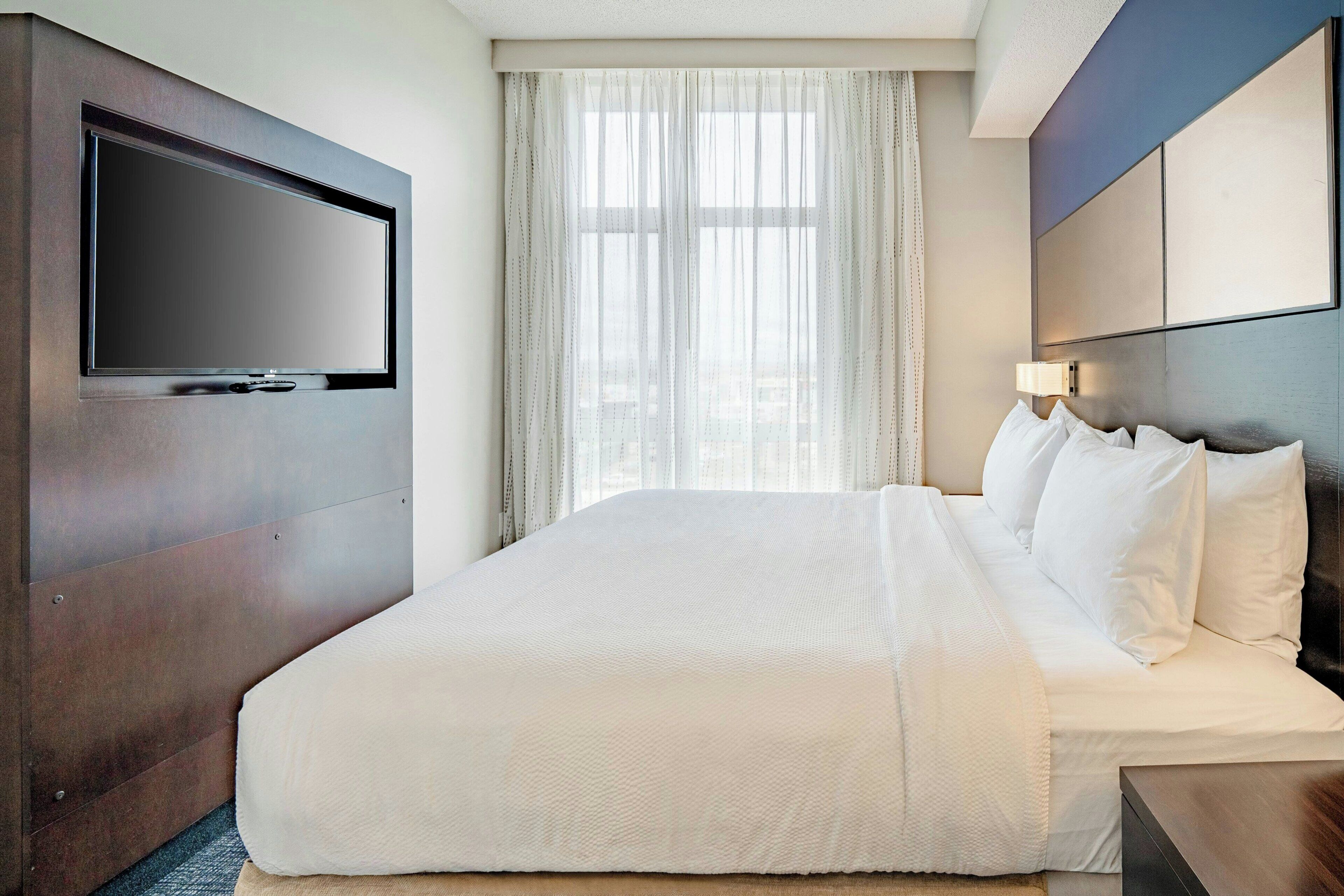Quarto de hotel com uma cama grande, televisão de tela plana fixada na parede, janela com cortinas translúcidas e abajur.