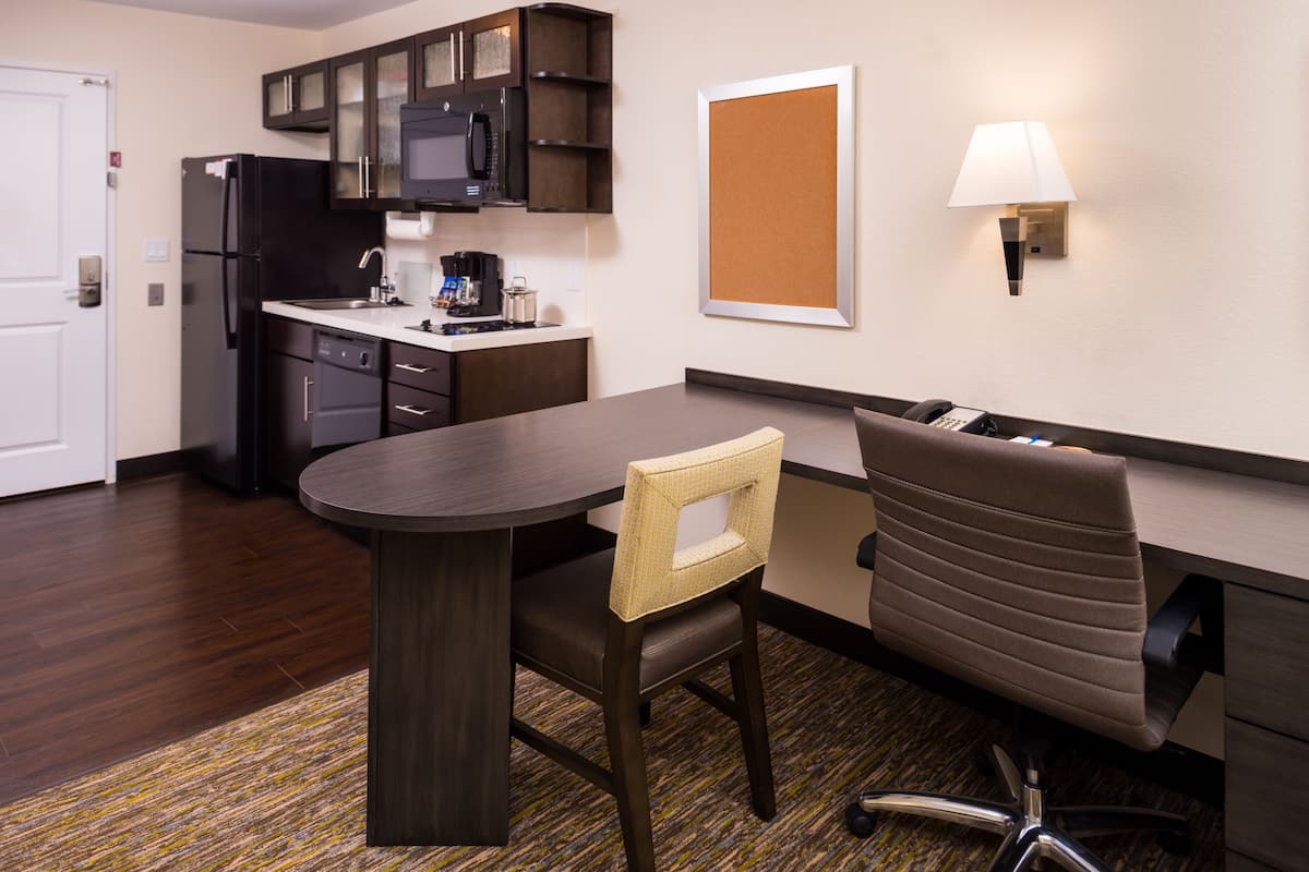 Quarto de hotel compacto com cozinha americana, mesa de jantar, uma cadeira, escrivaninha com cadeira e luminária fixada na parede.