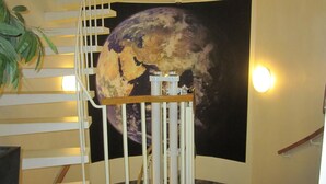 Uma escadaria em espiral com um mural da Terra ao fundo, uma planta à esquerda e um candeeiro fixado na parede à direita.