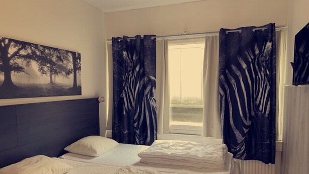 Quarto de hotel com duas camas, cabeceira, janela com cortinas e um quadro na parede.