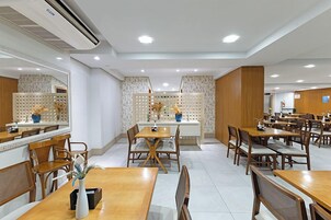 Uma ampla área de jantar com várias mesas e cadeiras, uma parede decorativa central e um aparelho de ar condicionado.