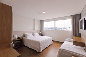 Quarto de hotel com uma cama grande, uma cama menor, uma escrivaninha, uma cadeira, uma televisão e uma janela com vista para a cidade.