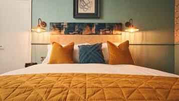Uma cama com cobertura acolchoada, duas luminárias fixadas na parede e uma obra de arte emoldurada acima da cama.