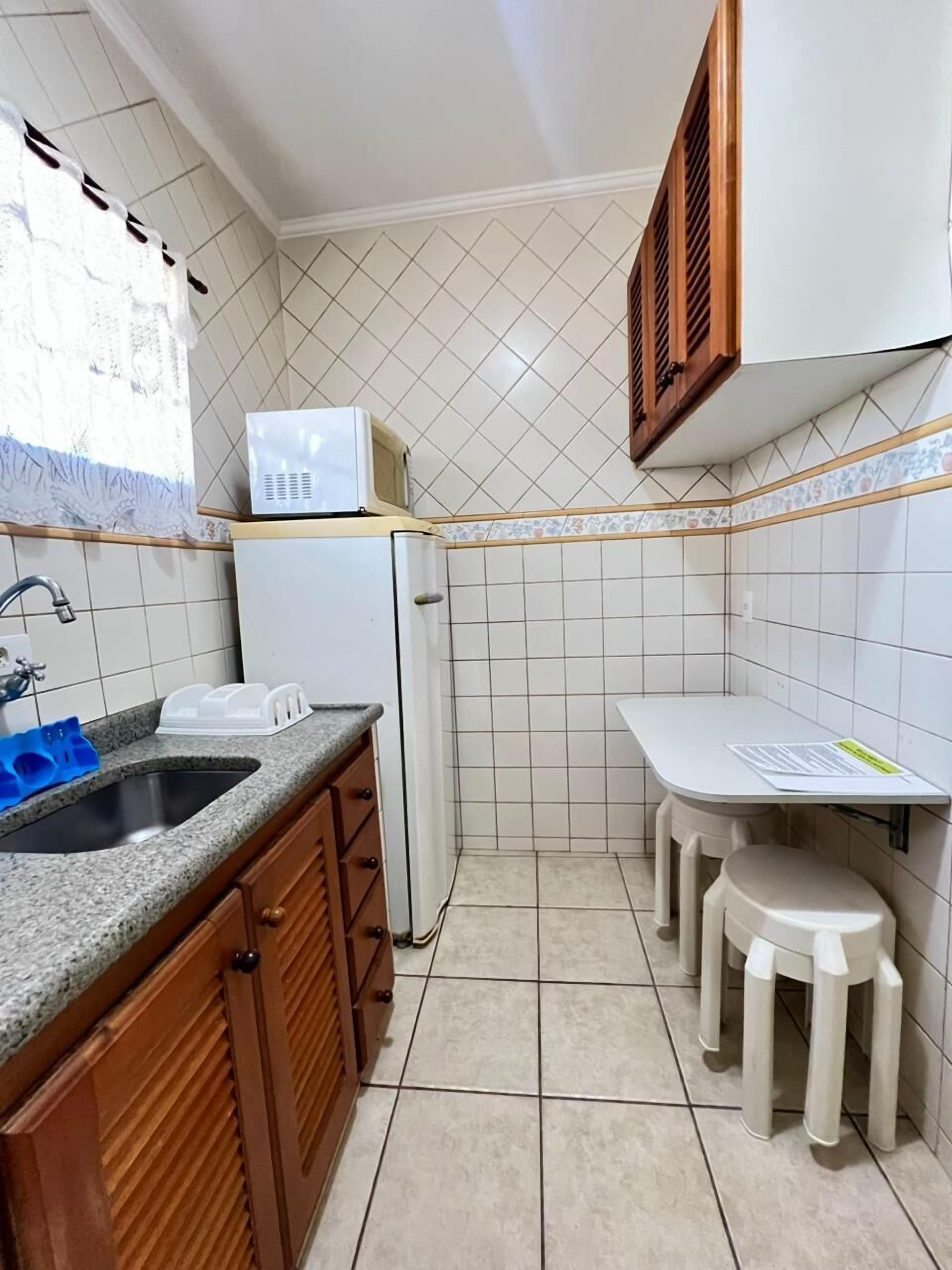 Uma cozinha compacta com pia, micro-ondas, geladeira, armários de madeira e um banquinho pequeno.