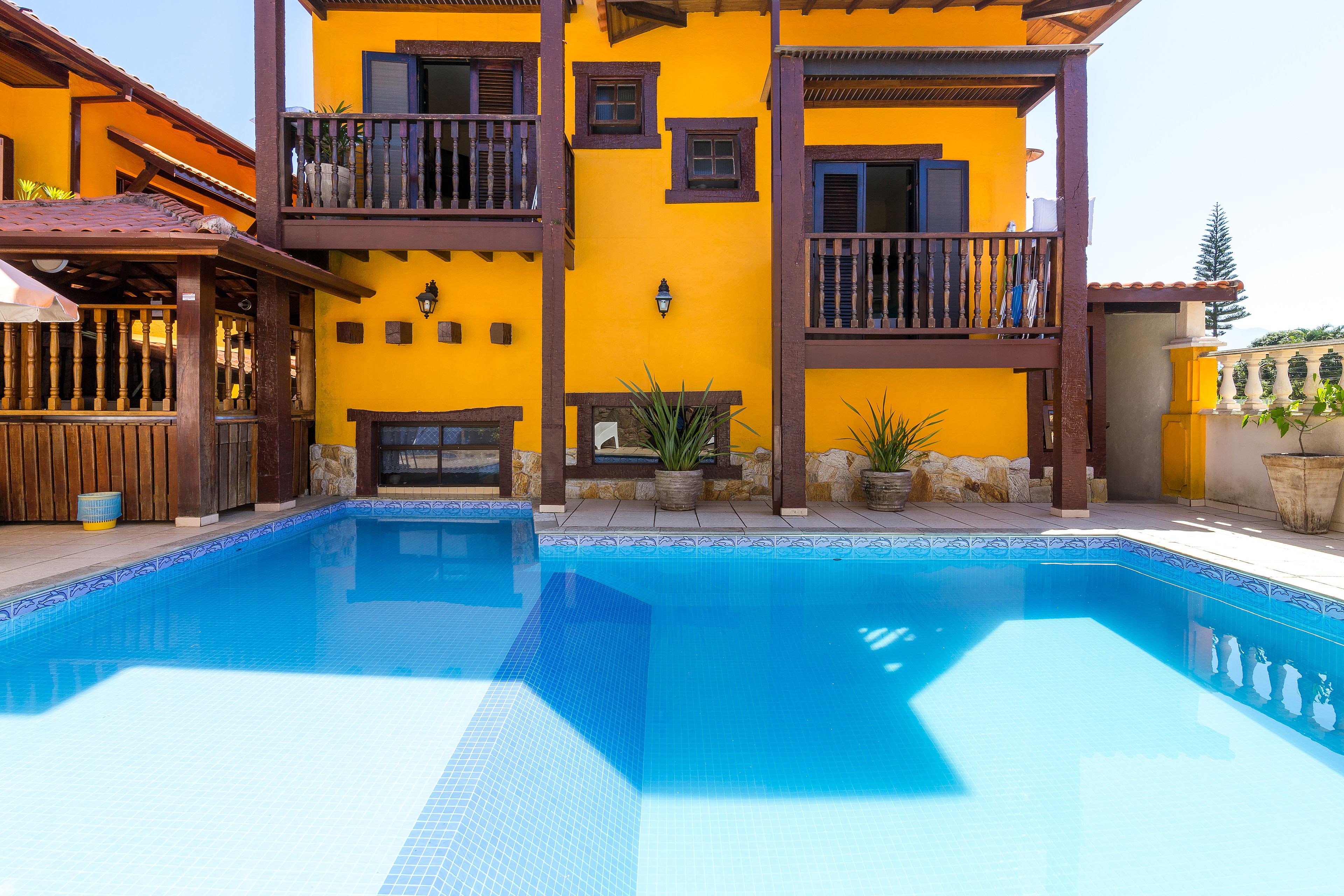 Piscina com borda azulejada, deck de madeira e edifício amarelo com varandas.
