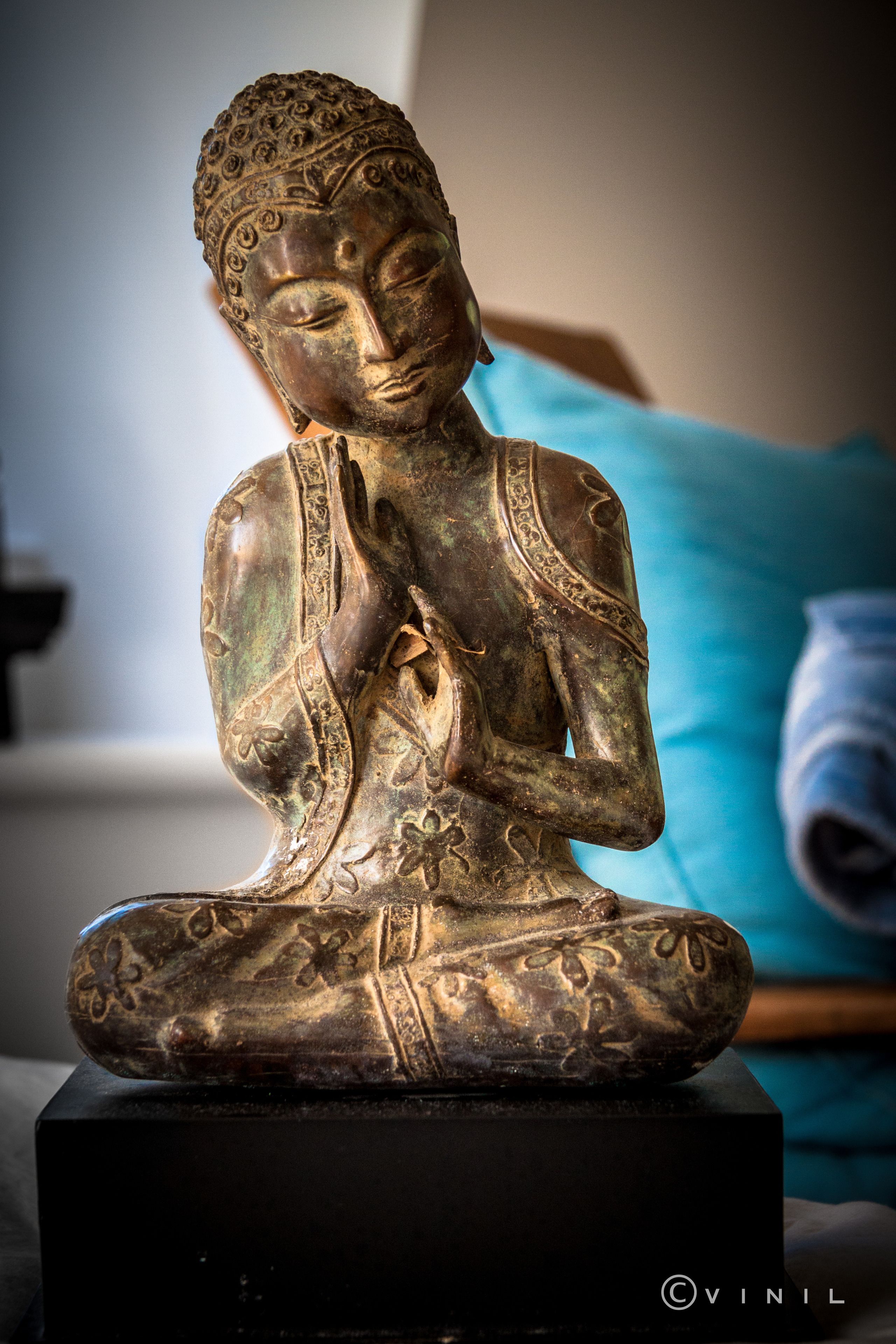 Uma estátua de Buda em bronze sobre um pedestal, numa sala com uma almofada azul e um tecido branco ao fundo.