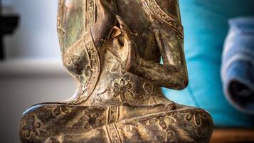 Uma estátua de Buda em bronze sobre um pedestal, numa sala com uma almofada azul e um tecido branco ao fundo.