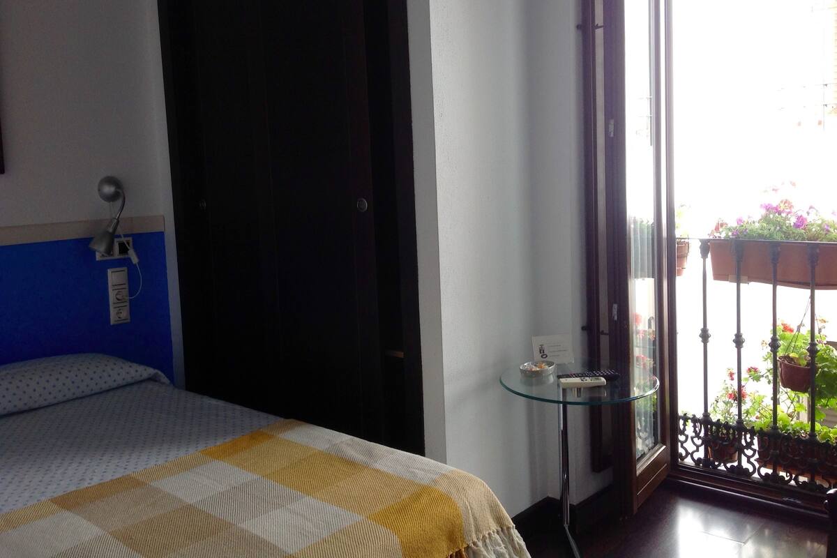 Quarto com cama, mesa de cabeceira com abajur, tomada e janela com vista para uma varanda com plantas em vasos.