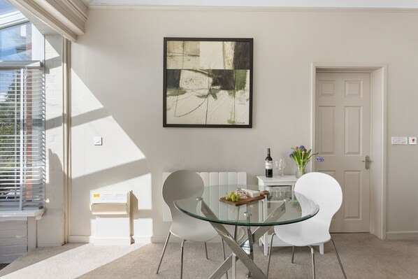 Uma área de refeições moderna com mesa de vidro, cadeiras brancas, um quadro abstrato emoldurado e uma porta à direita.