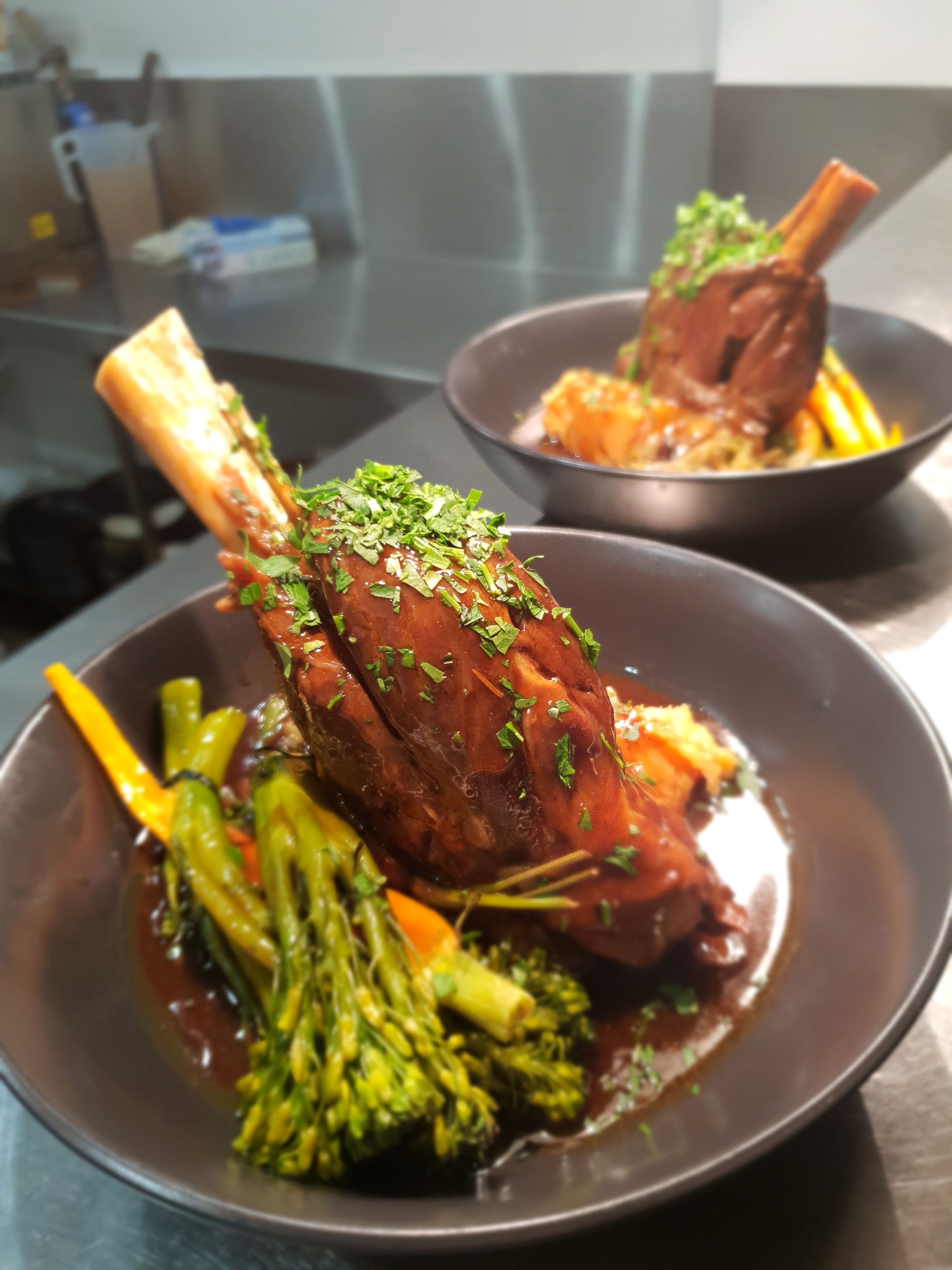 Prato com um ossobuco de cordeiro assado, legumes verdes e molho escuro.