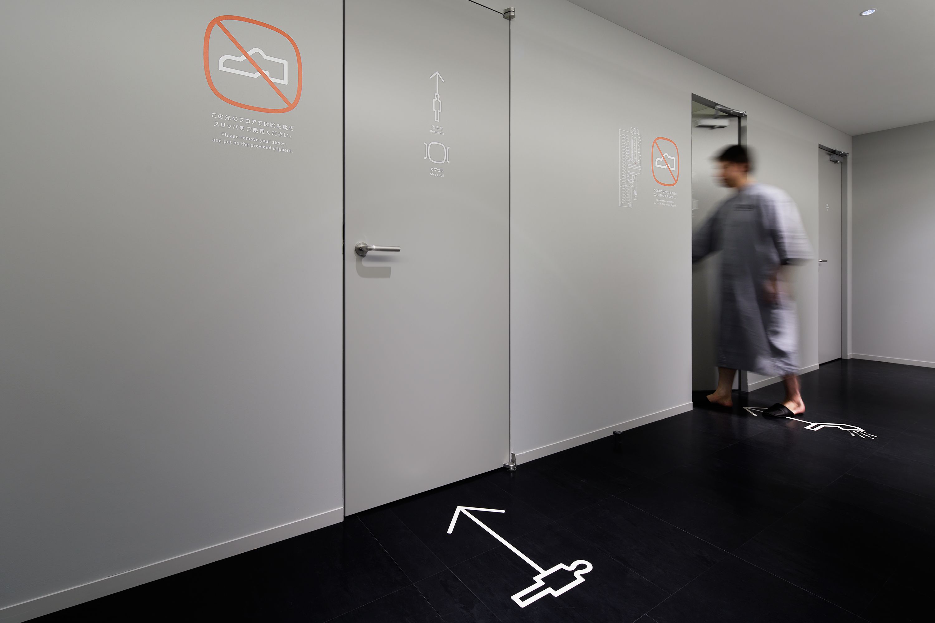 Um corredor moderno com setas indicativas no chão, uma pessoa caminhando e uma porta com sinal de proibido fumar.