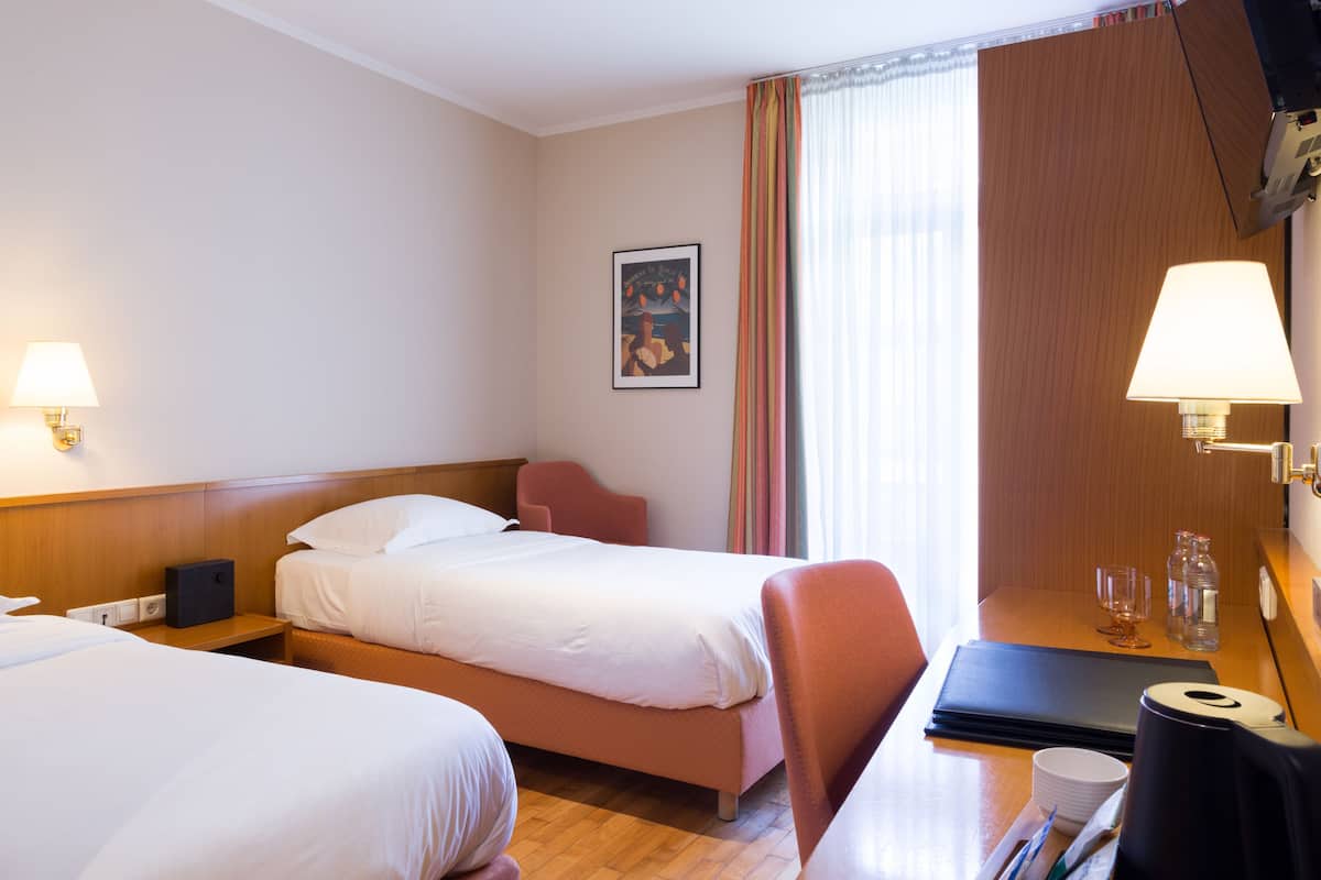 Quarto de hotel com duas camas, uma escrivaninha com abajur, um quadro na parede e uma janela com cortinas.