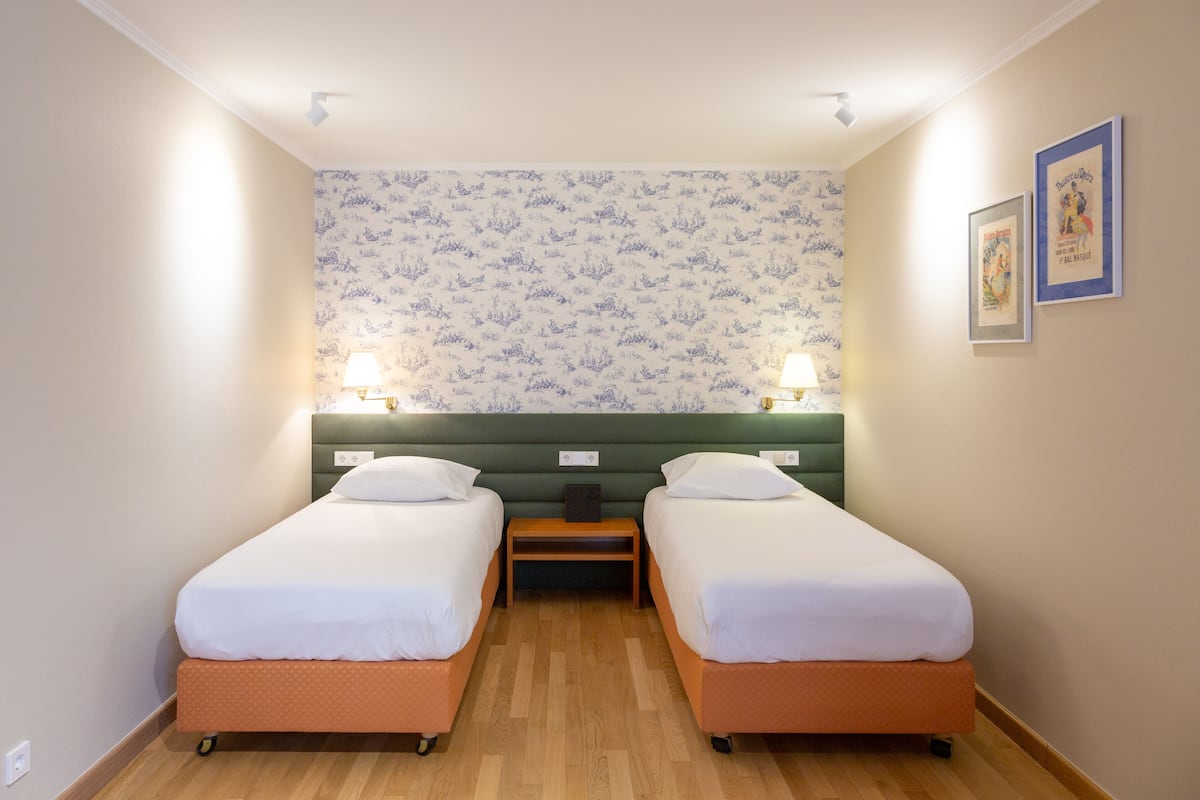 Quarto de hotel com duas camas de solteiro, cabeceira verde, papel de parede estampado com flores e dois quadros na parede.