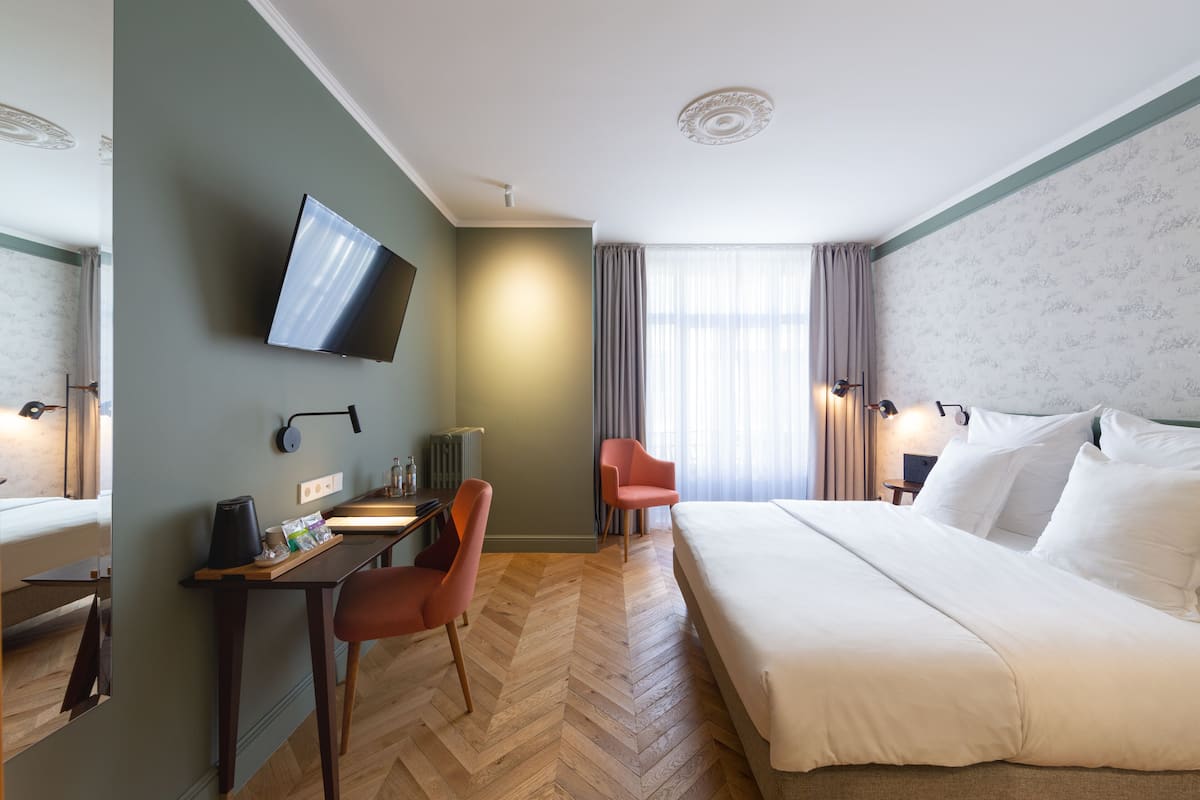 Quarto de hotel com uma cama, uma escrivaninha com cadeira, televisão e janela com cortinas.