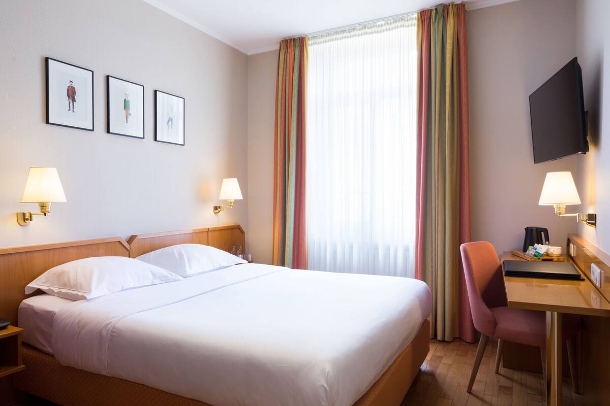 Quarto de hotel com uma cama grande, duas mesas de cabeceira com abajures, uma escrivaninha com cadeira, televisão de tela plana e uma janela com cortinas.