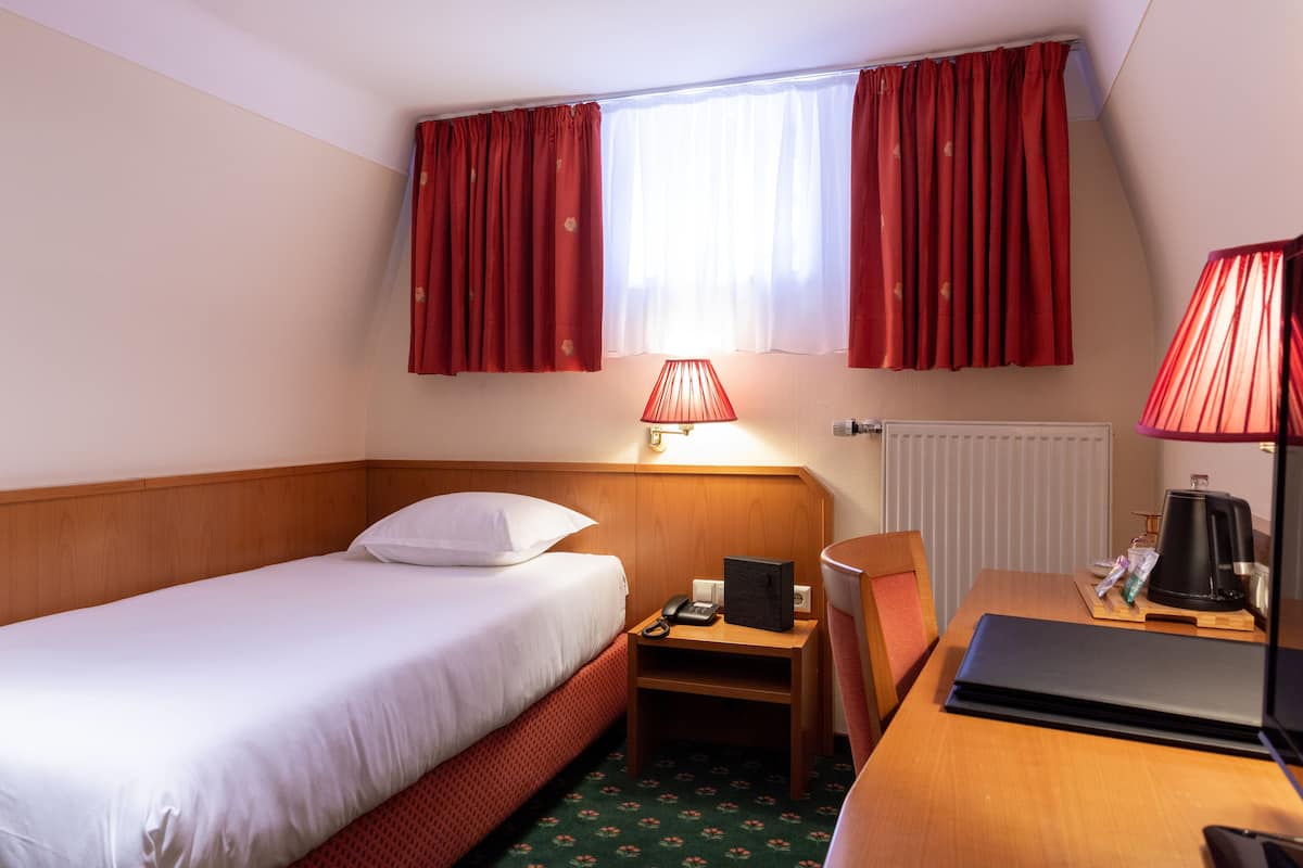Quarto de hotel com uma cama de solteiro, uma escrivaninha com luminária, uma cadeira, uma cafeteira e uma janela com cortinas vermelhas.