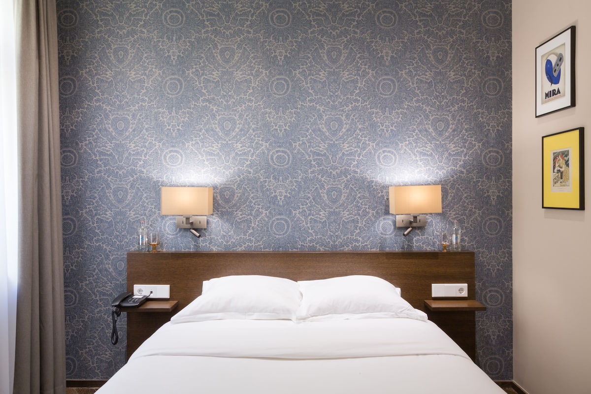 Quarto de hotel com papel de parede estampado, duas luminárias de cabeceira, uma cabeceira de madeira, um telefone ao lado da cama e um quadro na parede.