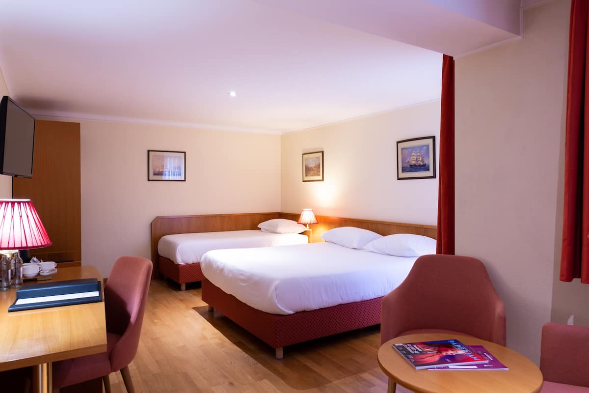 Quarto de hotel com duas camas, uma escrivaninha com abajur, uma cadeira e uma mesinha de centro com uma revista.